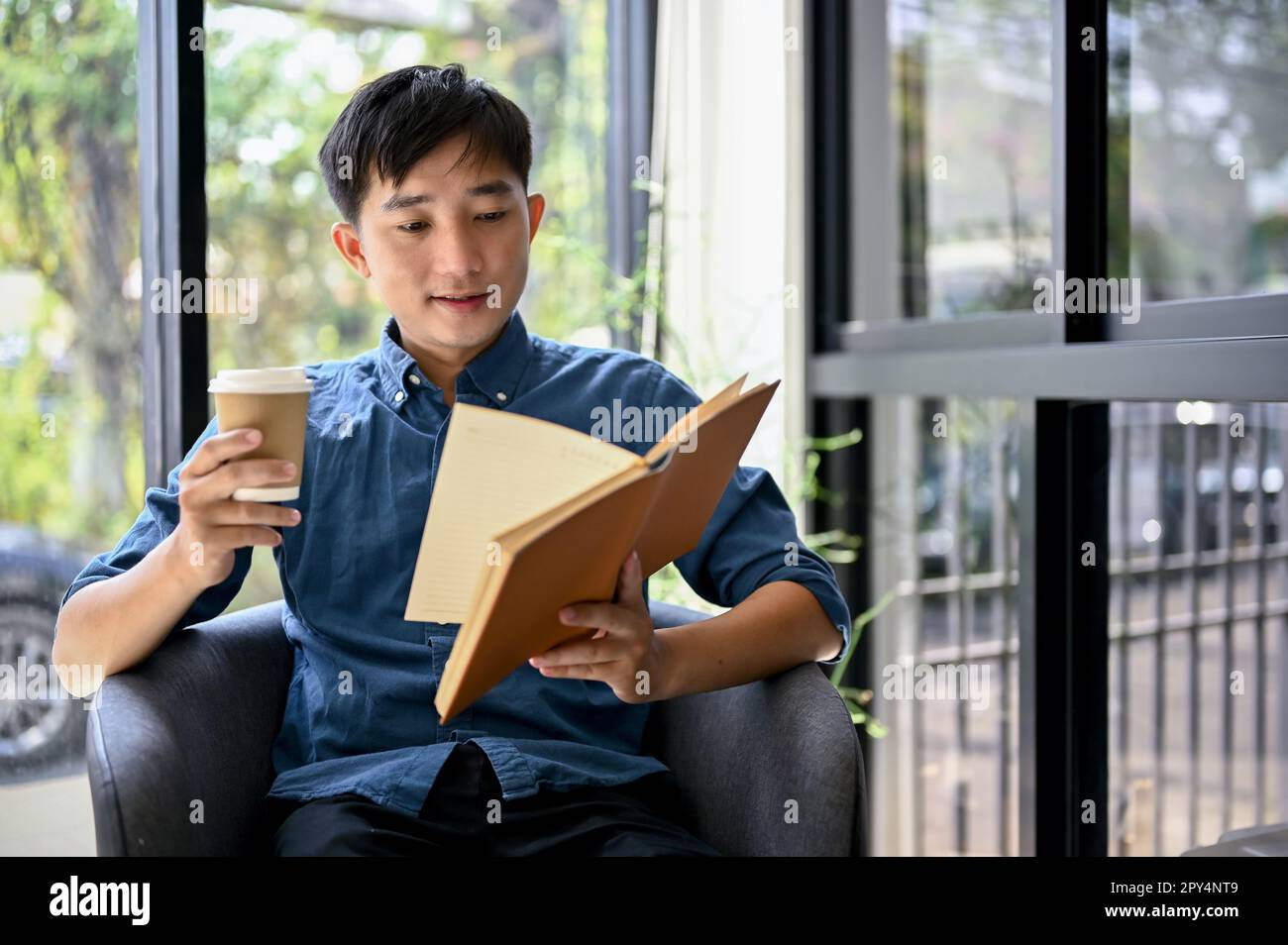 Happy millennial Asian man enjoying with his coffee and reading a book ...