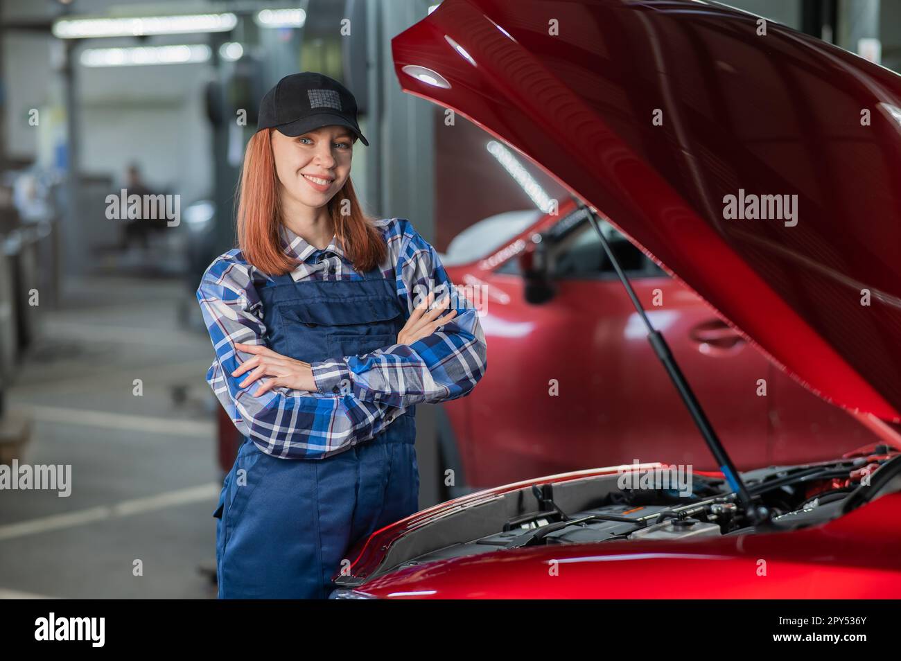 A woman auto mechanic stands at the open hood of a car with her arms ...