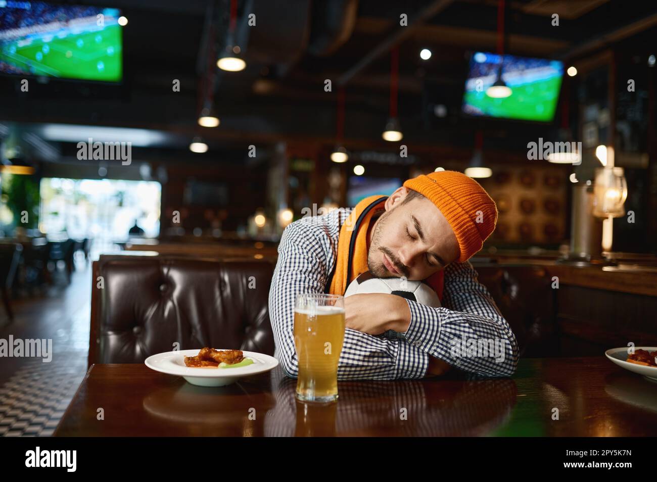 Funny young man soccer fan sleeping on ball while rest in sport bar ...