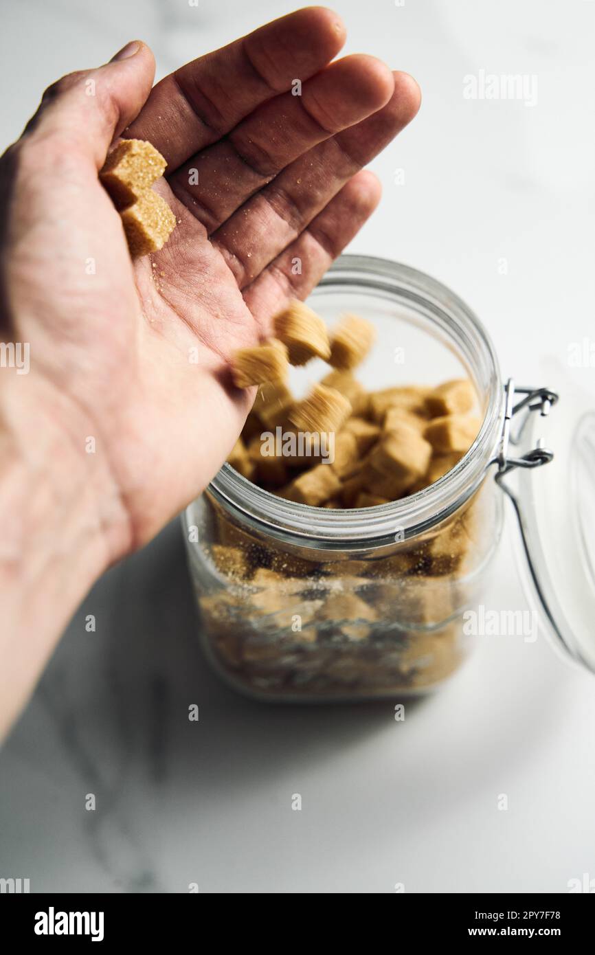 Brown sugar cubes in a man's hand and in the jar on white marble ...