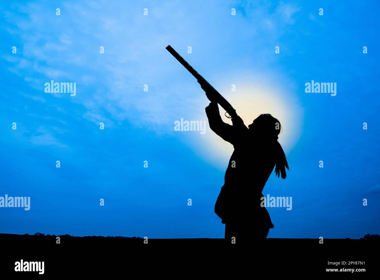 A silhouette portrait of a woman with a shotgun against a blue sky ...