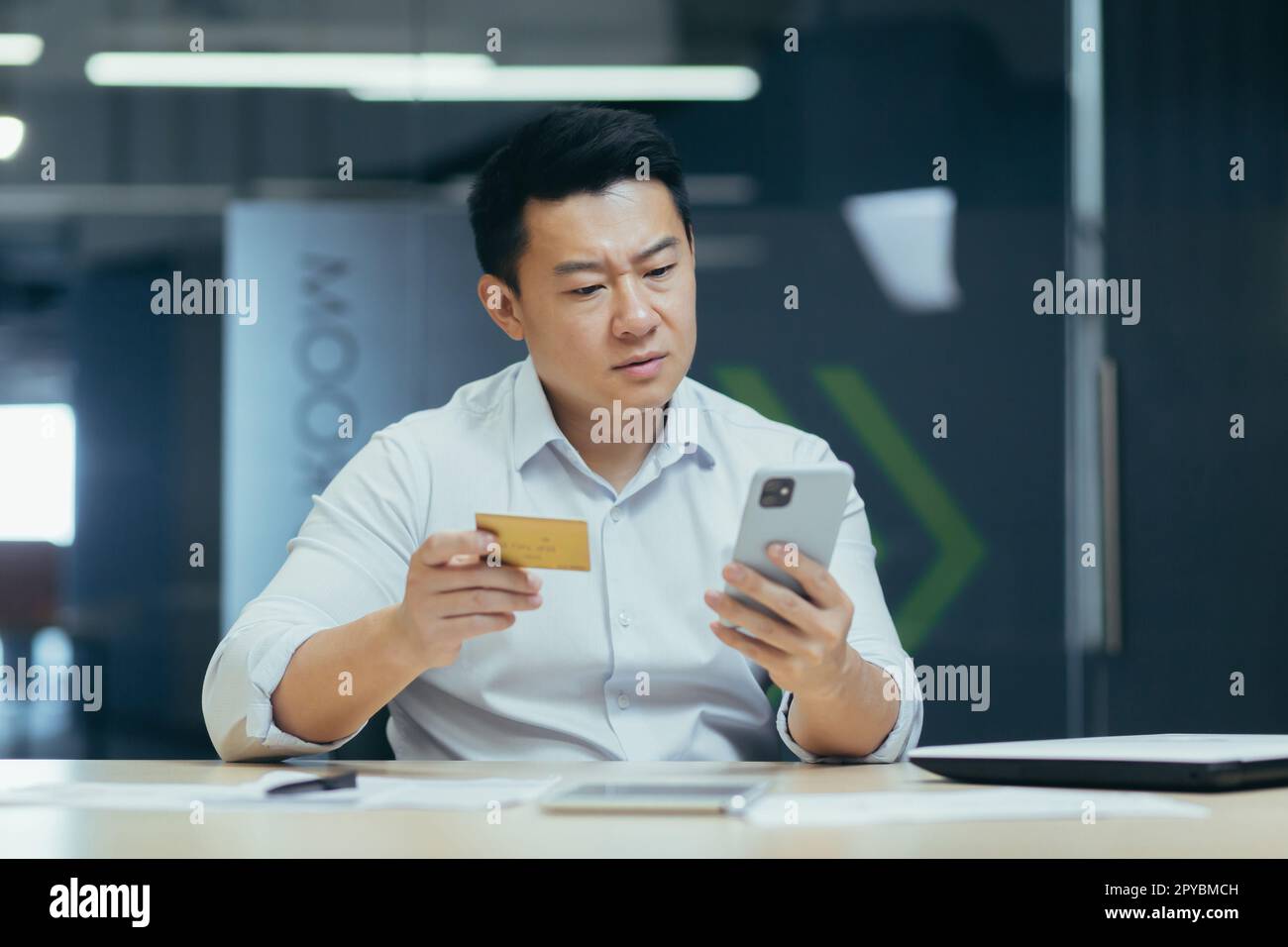 Worried young asian man sitting at desk in office and holding phone and ...