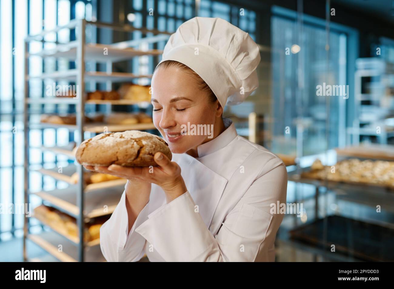 Young female baker smelling fresh bread over modern bakery background ...