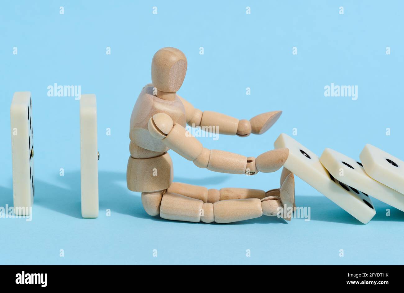 A wooden puppet toy holds back a falling domino on a blue background ...