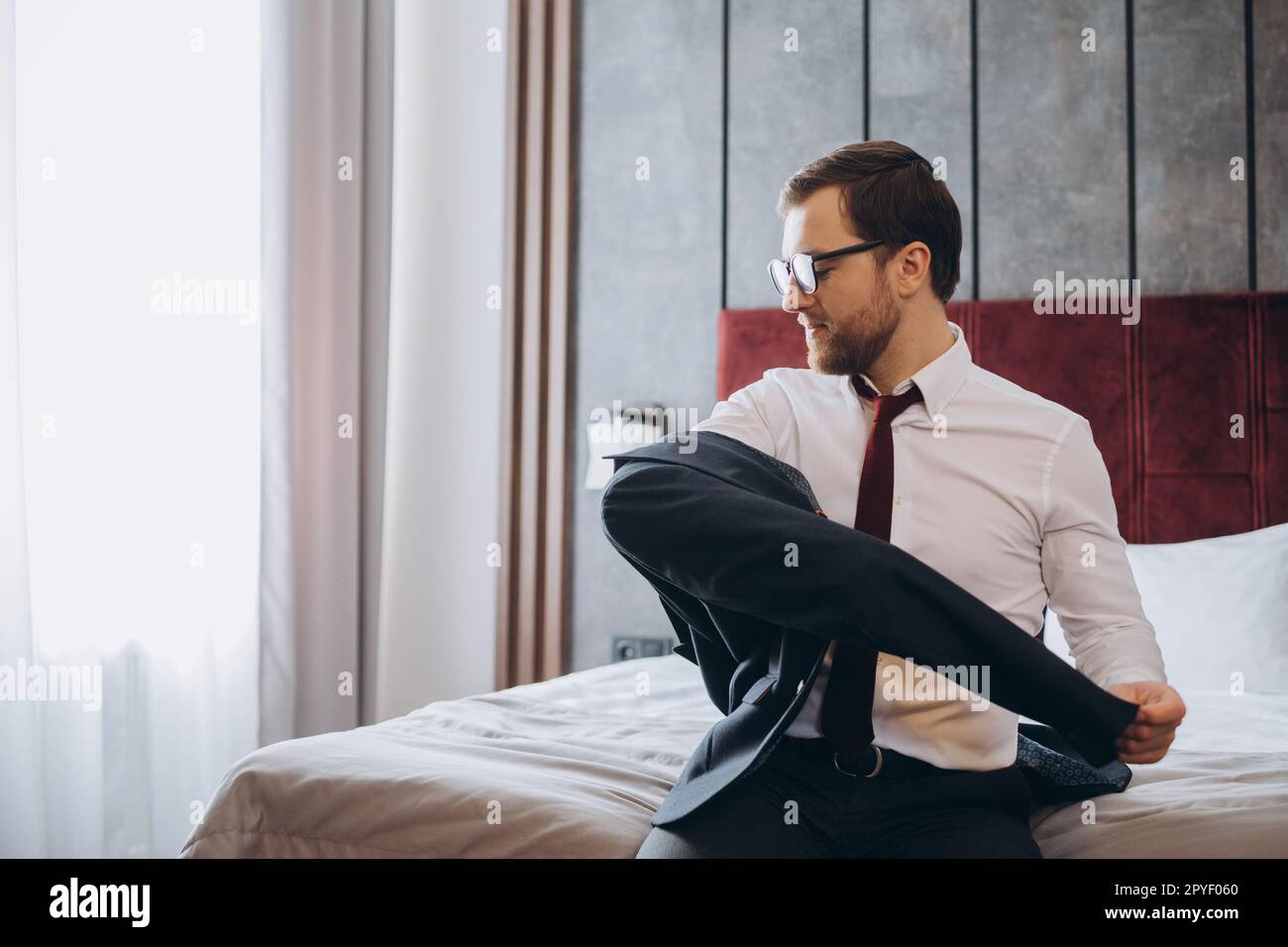Business man with suitcase taking off jacket while sitting on bed in ...
