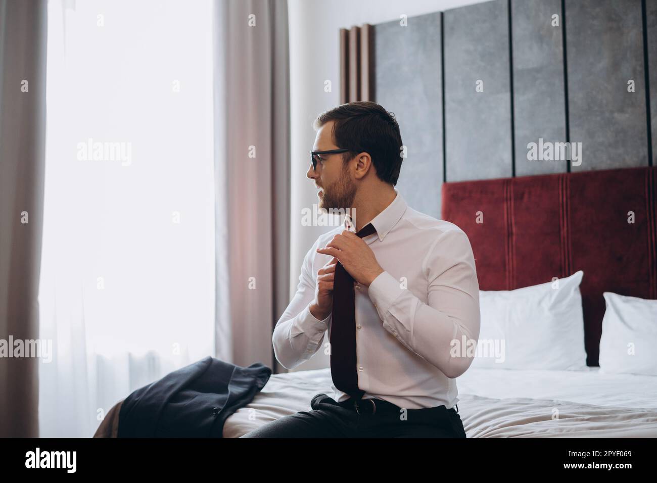 Young businessman taking off his tie while sitting on a bed in a hotel ...