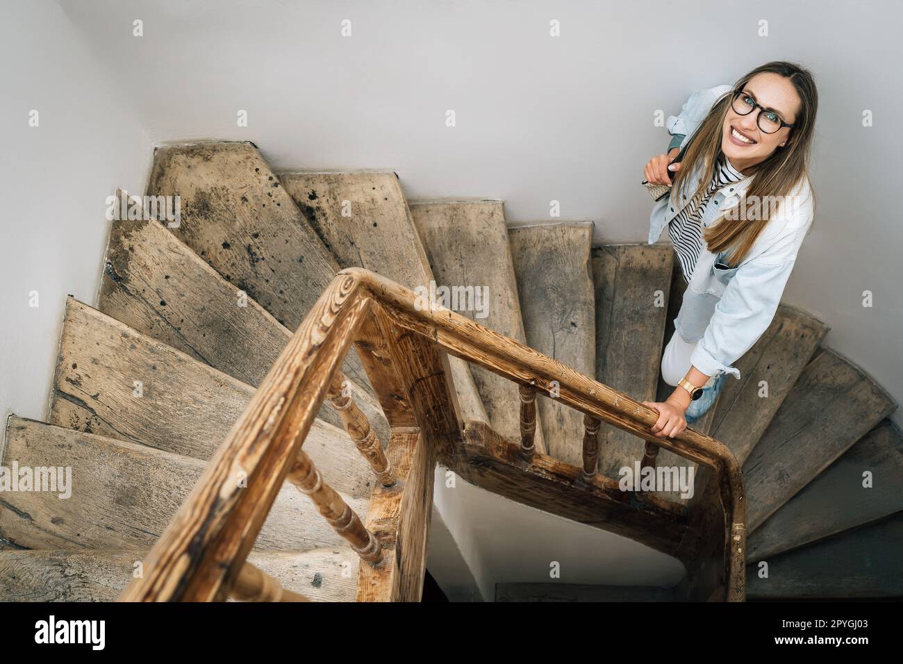 A woman with glasses smiling looks up climbing an old wooden staircase ...
