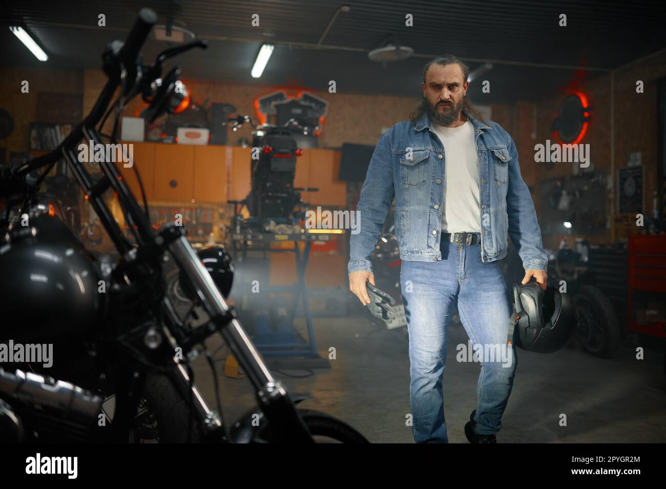 Portrait of bearded brutal biker looking motorcycle in garage workshop ...
