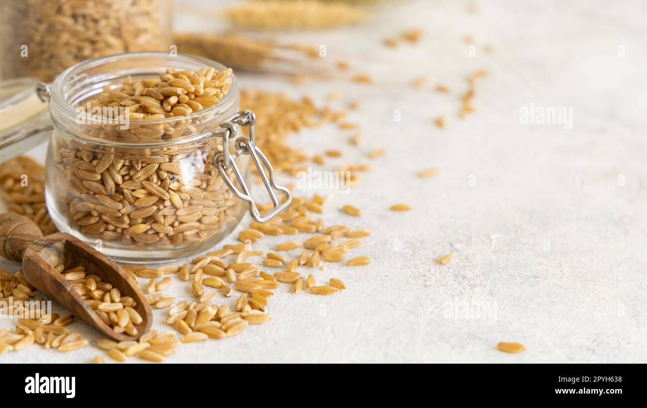 Glass jar of raw dry rye grain with a wooden spoon on white table close ...