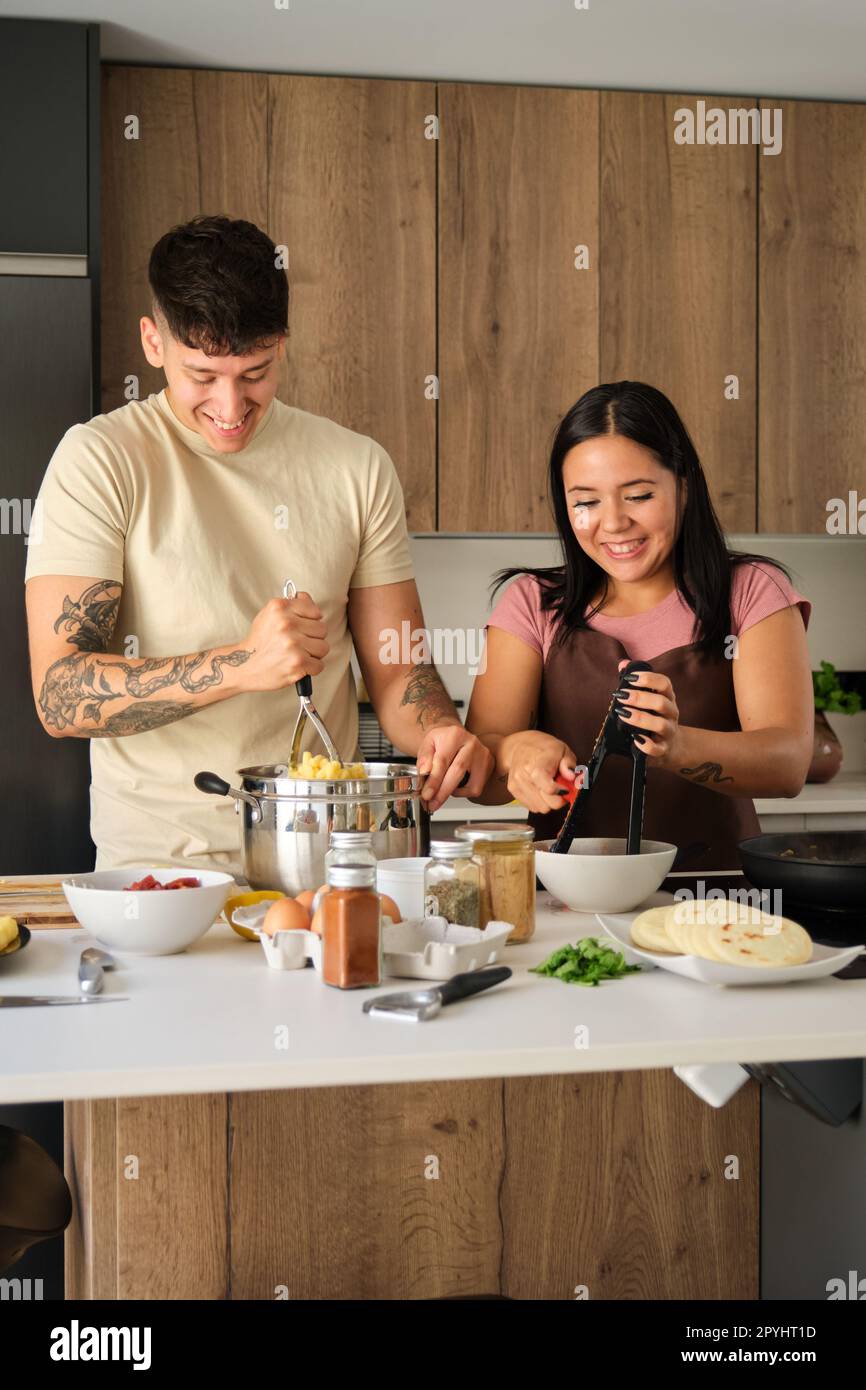 Two young latin people mashing potatoes and grating tomato at kitchen ...