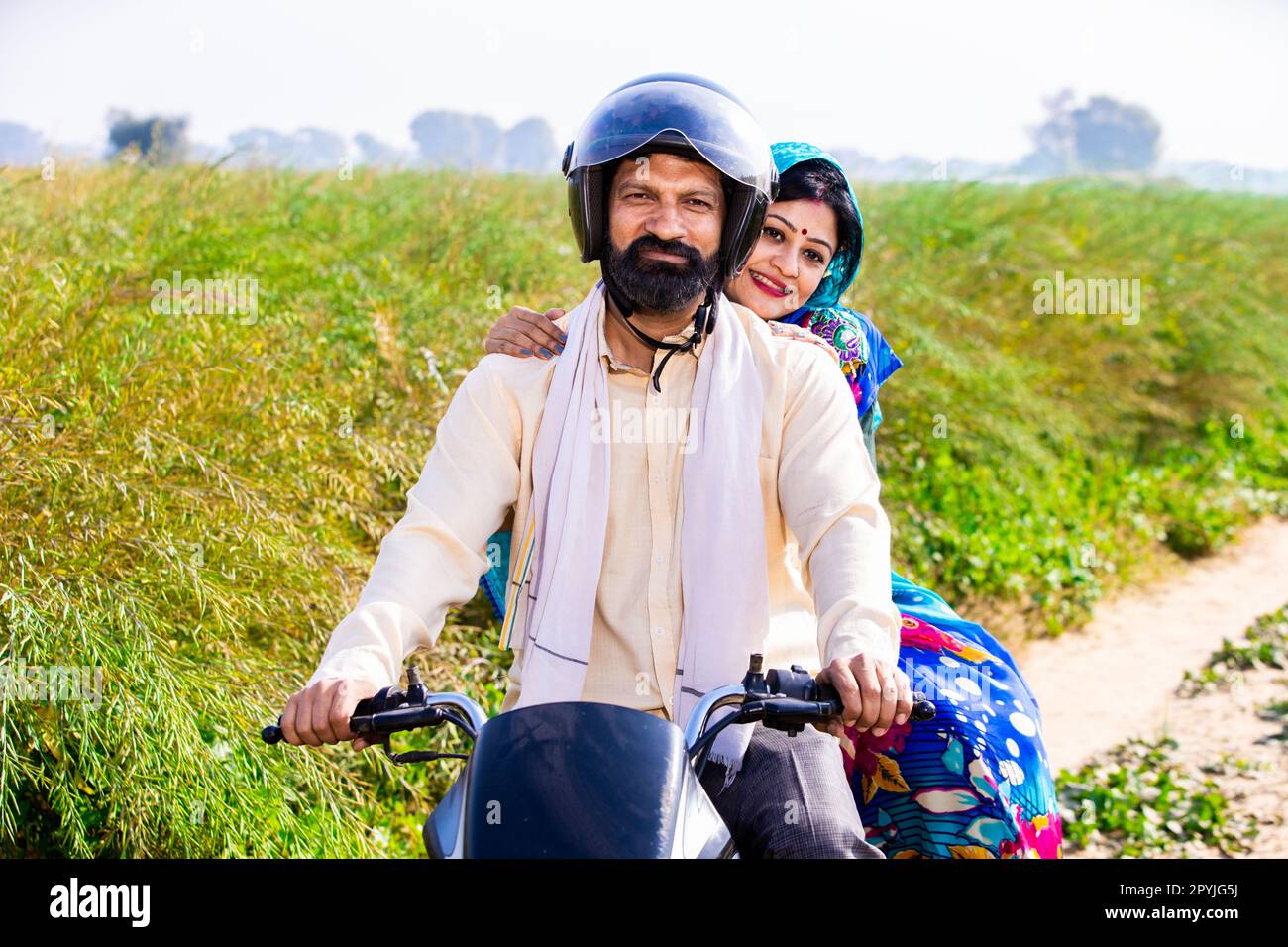 Happy young rural Indian couple wearing safety helmet riding on motorcycle in village ...