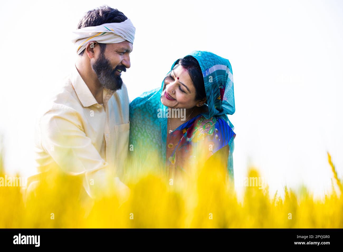Happy young indian farmer couple standing looking at each other at golden wheat crop agriculture ...