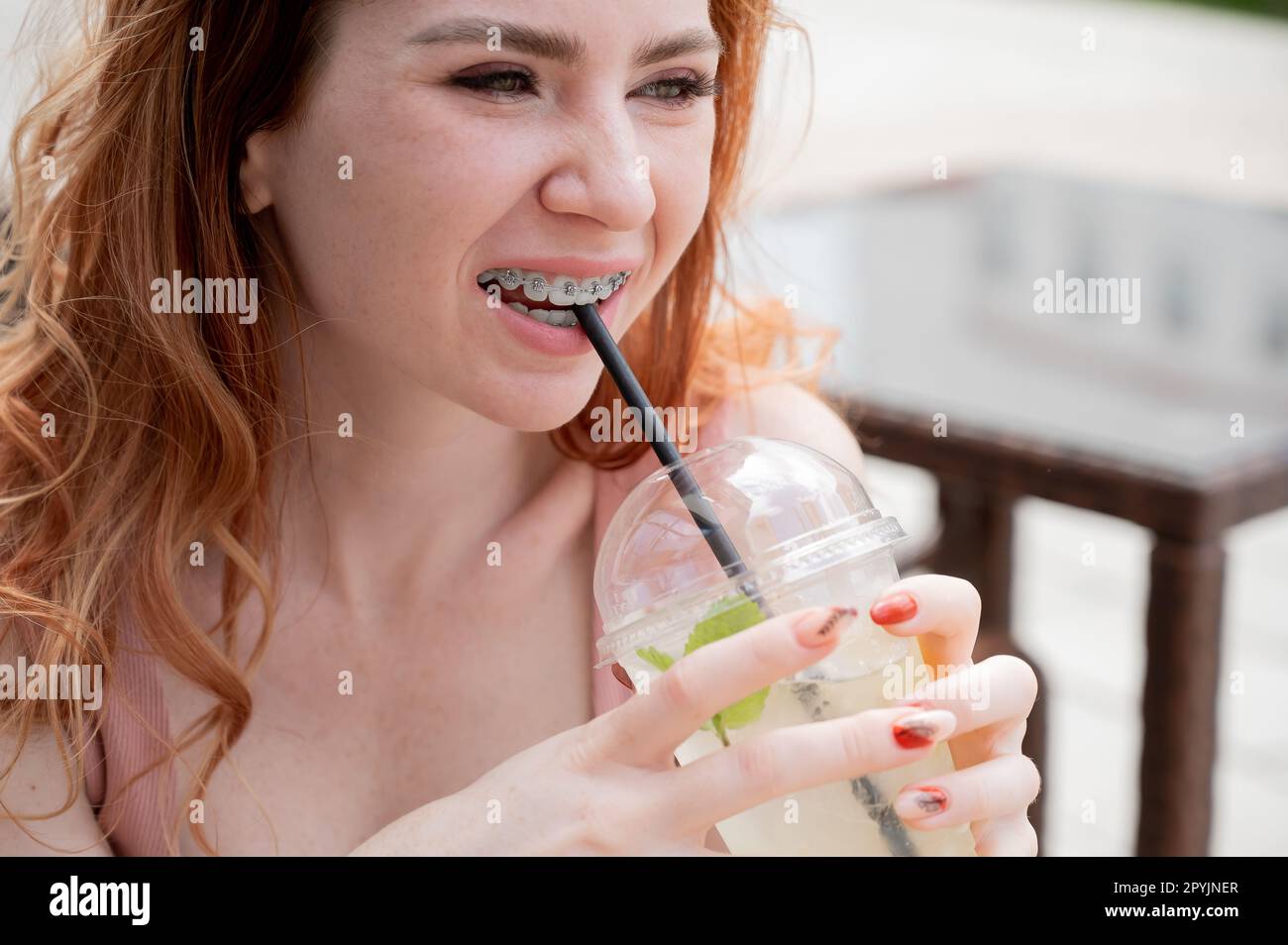 Young beautiful red-haired woman with braces drinks cooling lemonade ...