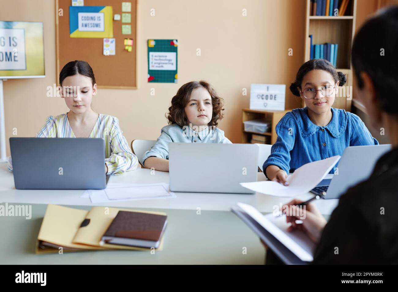 Group of school children sitting at table with computers and listening ...
