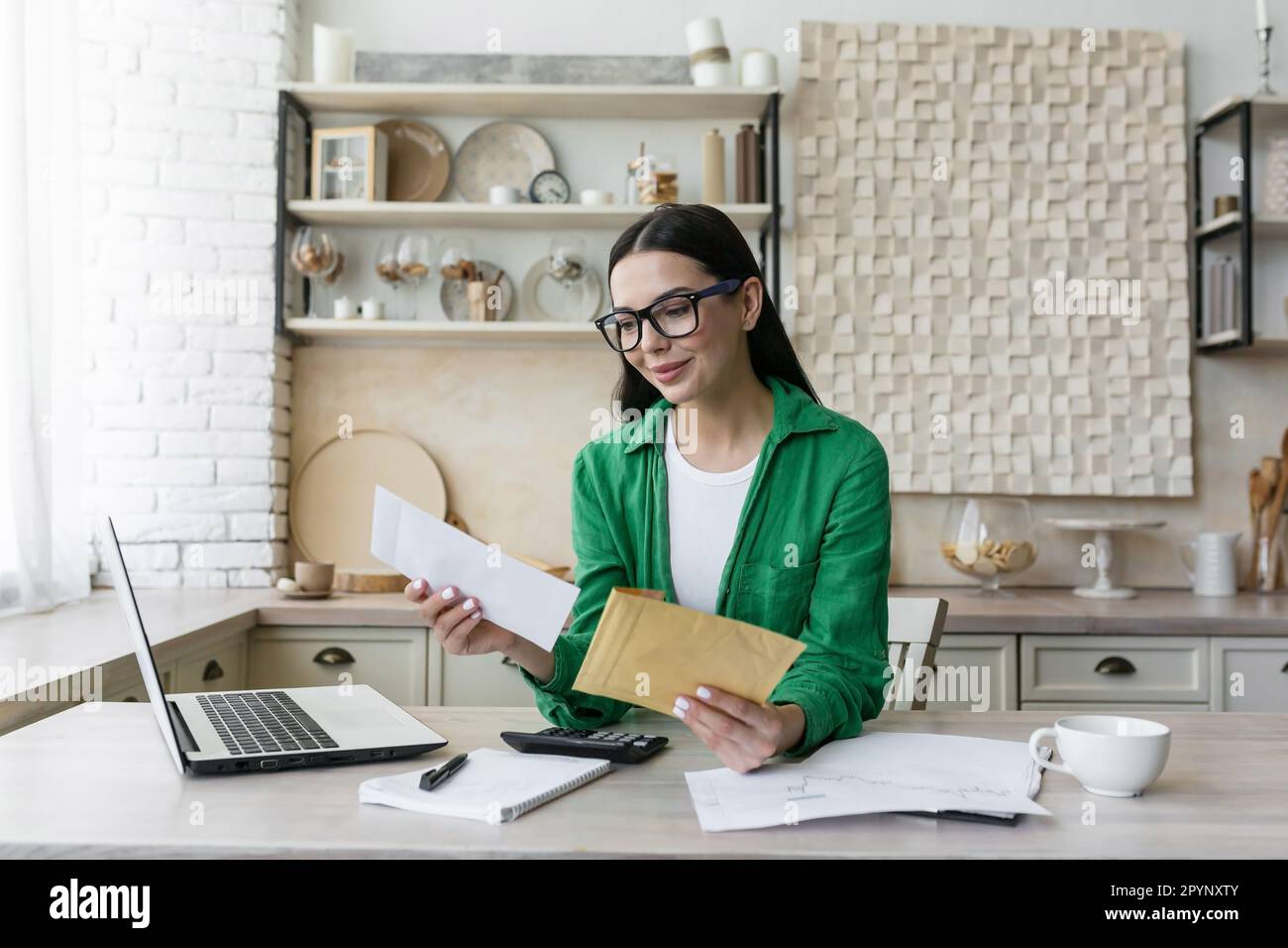 Smiling young woman working at home with laptop and documents. He opens the letter from the ...