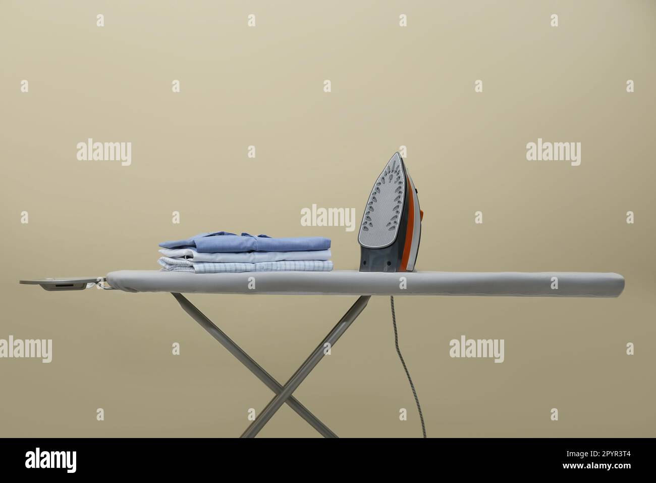 Modern iron and stack of clean clothes on board against beige ...
