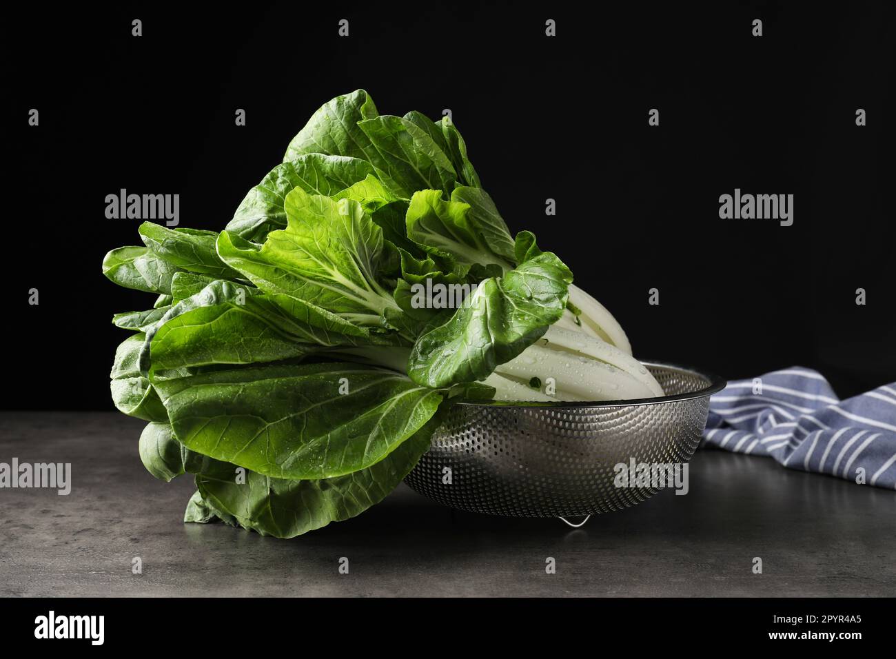 Fresh green pak choy cabbages with water drops in sieve on grey table ...