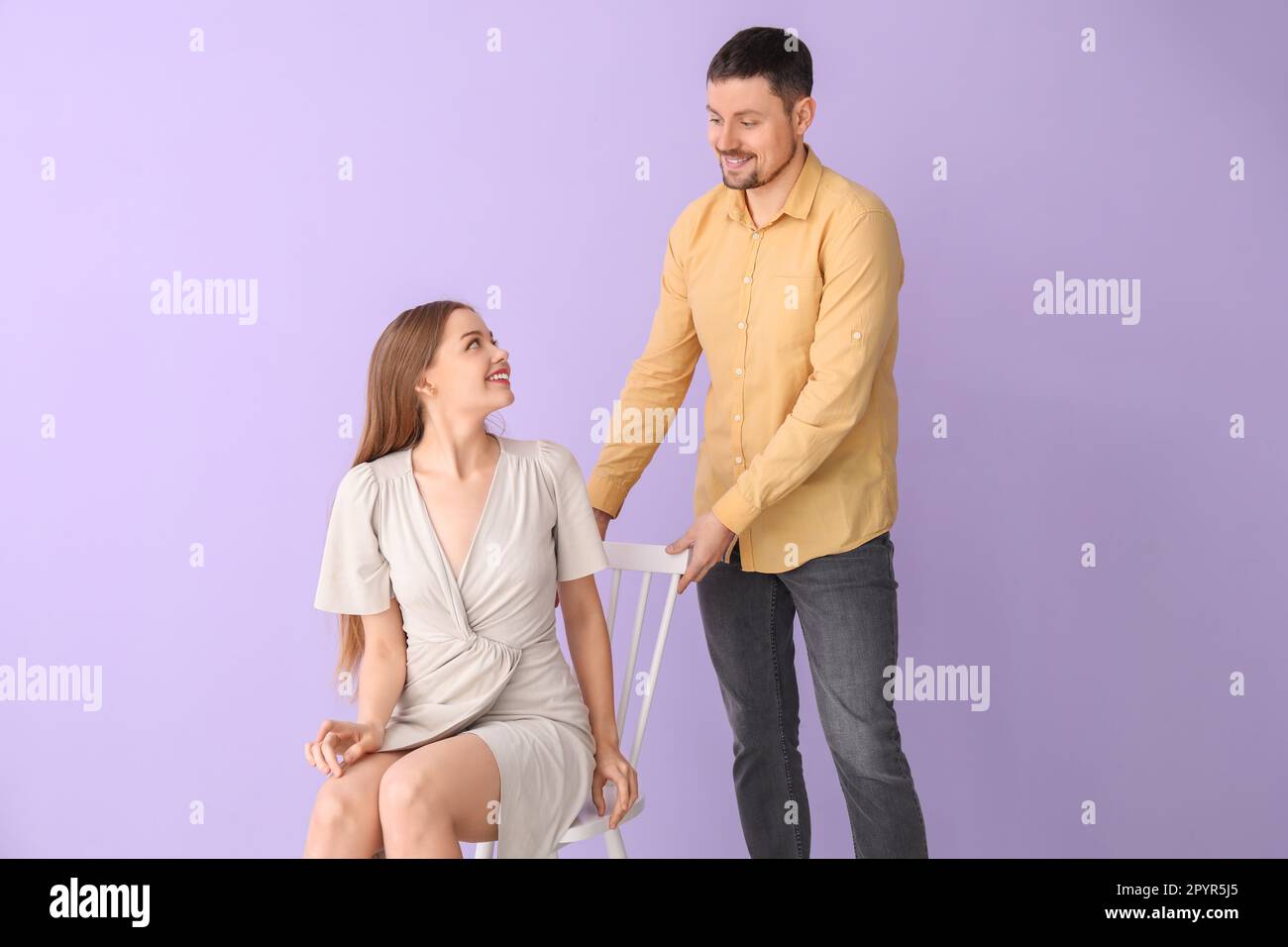 Young man pulling up chair for his wife on lilac background Stock Photo ...