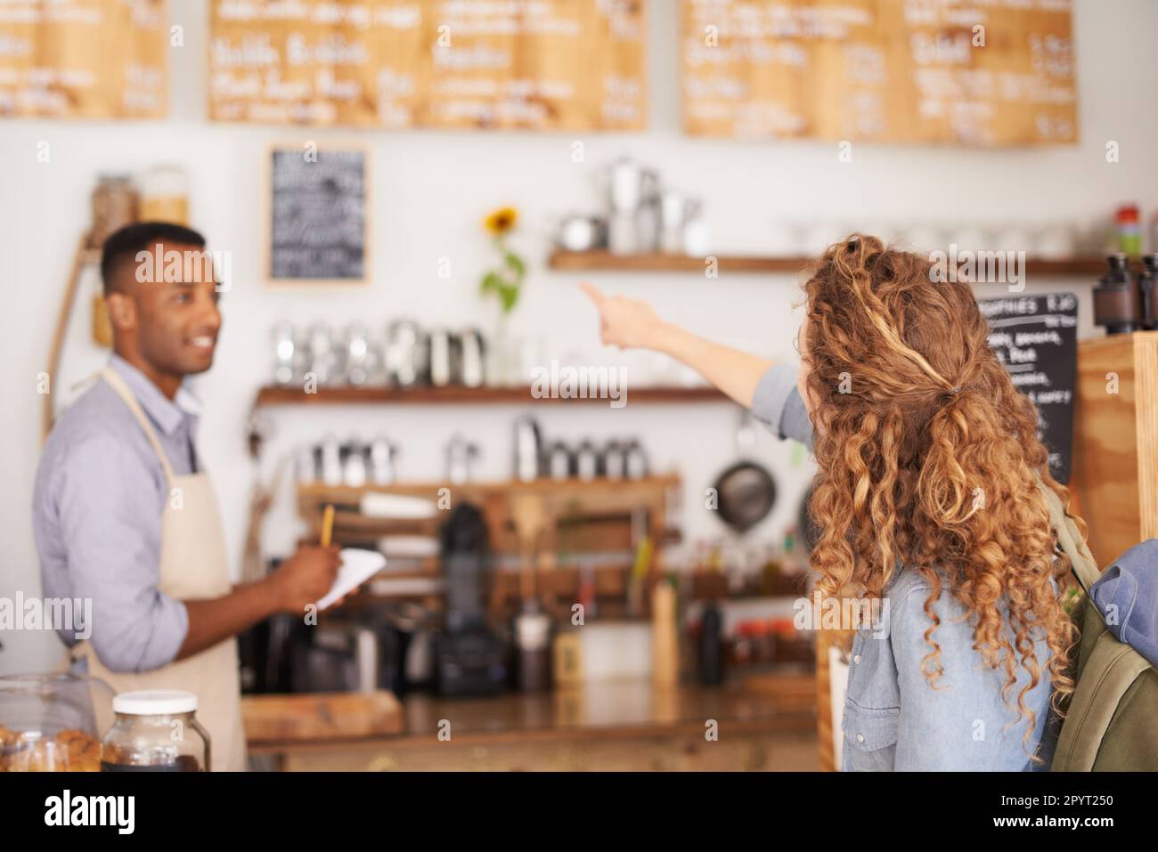 Woman, coffee shop and pointing at menu on wall with barista, notes and ...