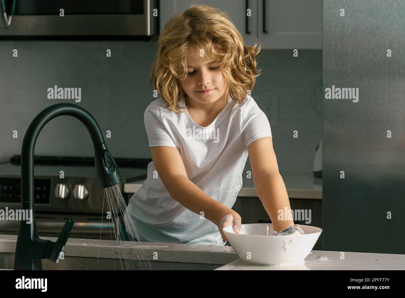 Kid boy washing dishes in the kitchen interior. Child helping his ...