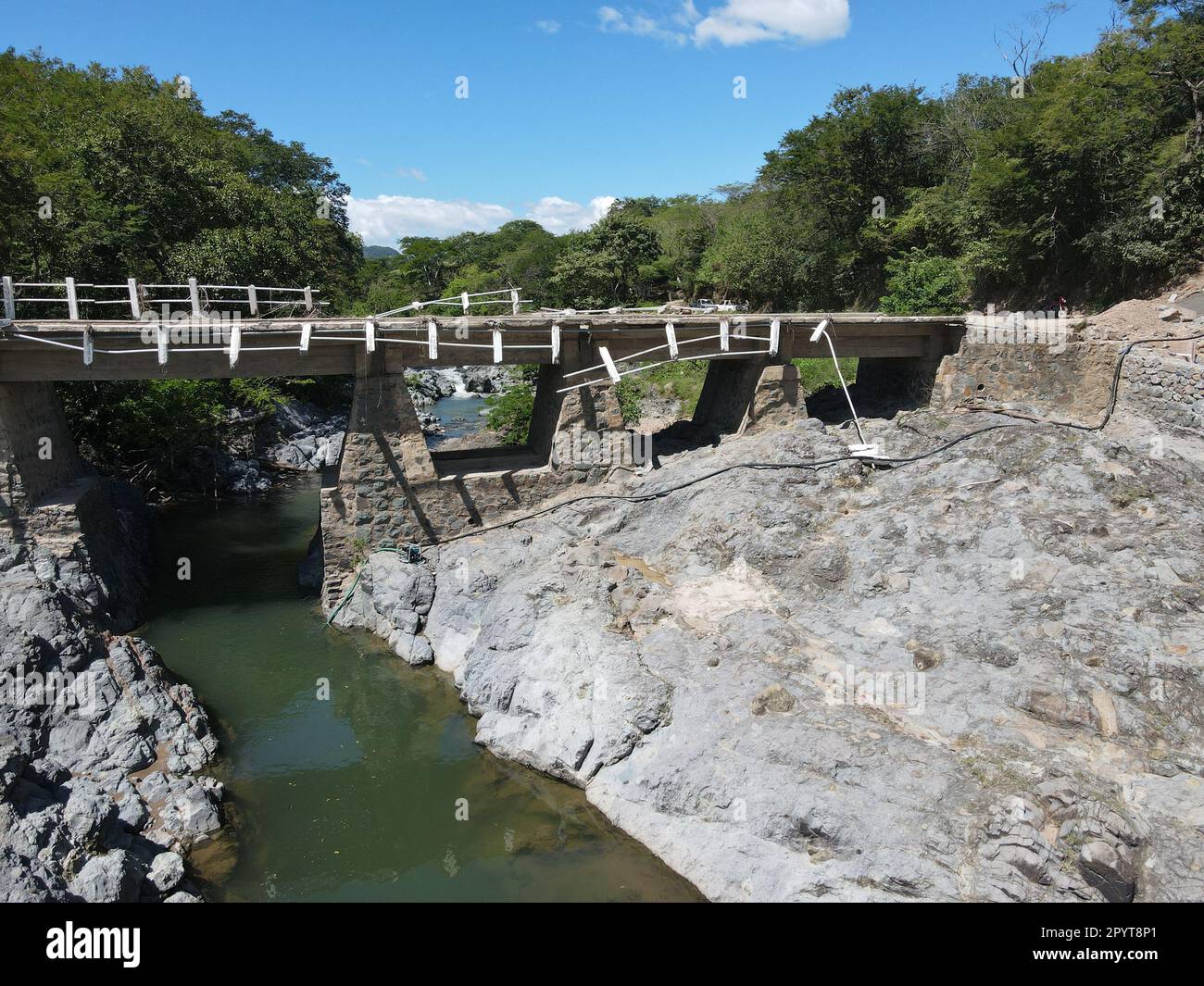 A damaged bridge over a river with large rocks and dark green water ...