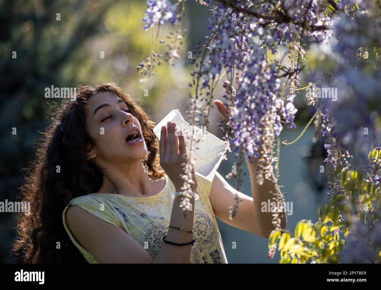 Pretty young woman sneezing and blowing nose in front of blue rain tree ...