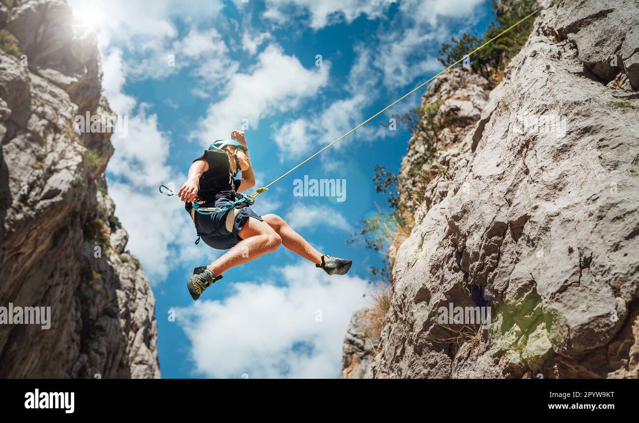 Climber teenage boy in protective helmet jumping on vertical cliff rock wall using rope Belay ...