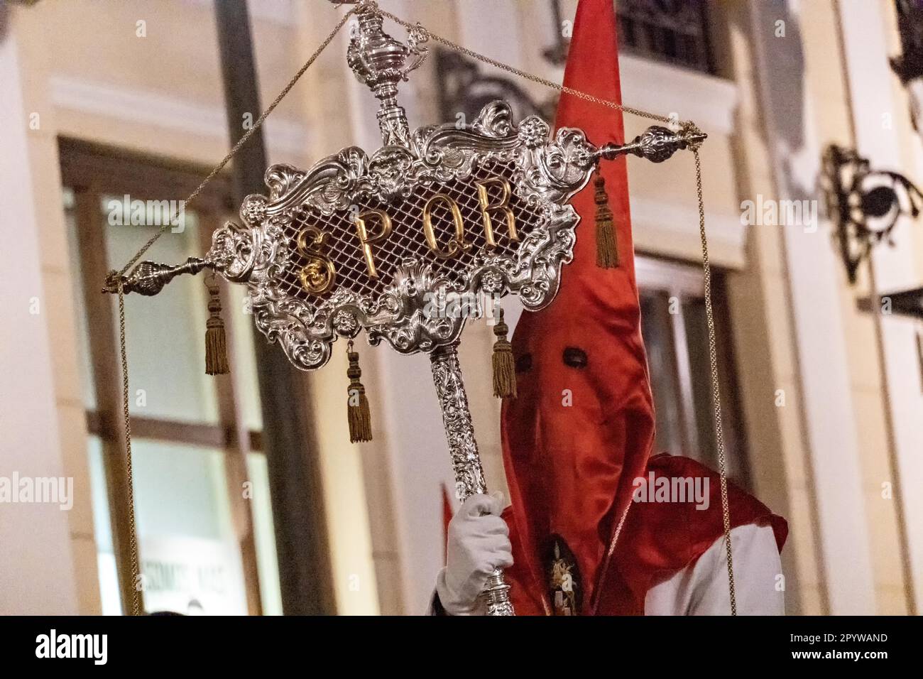 A Cofradia wearing a red cone shaped hood carries a sign with the ...