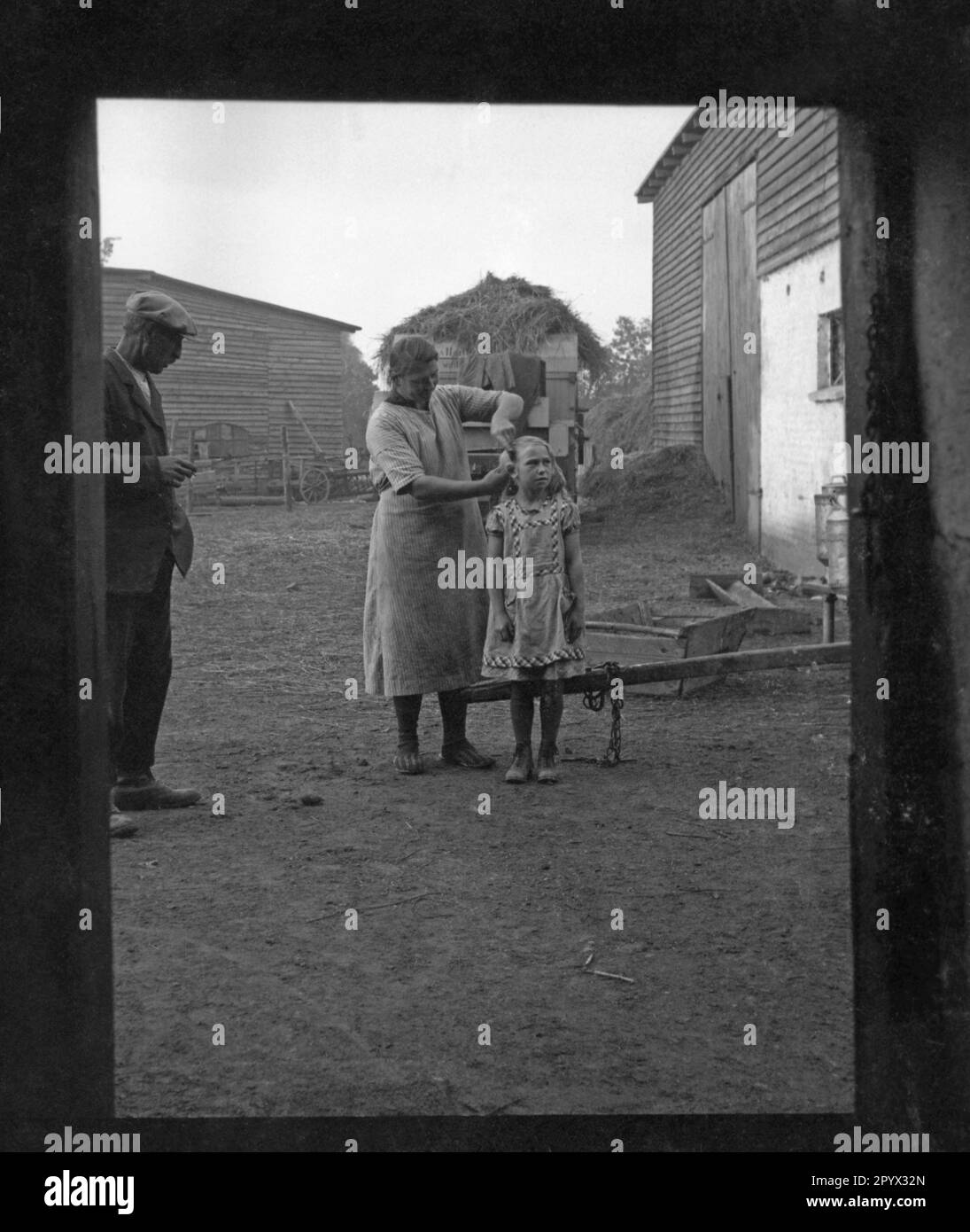 On a farm, a woman is braiding the hair of a girl wearing a traditional ...