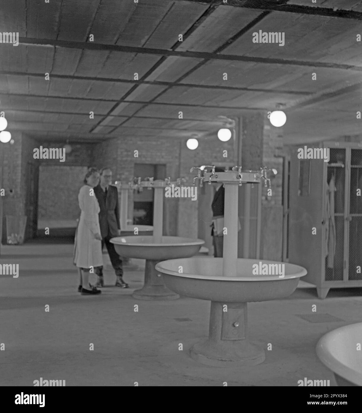 A man and a woman stand in a room with several large sinks and ...