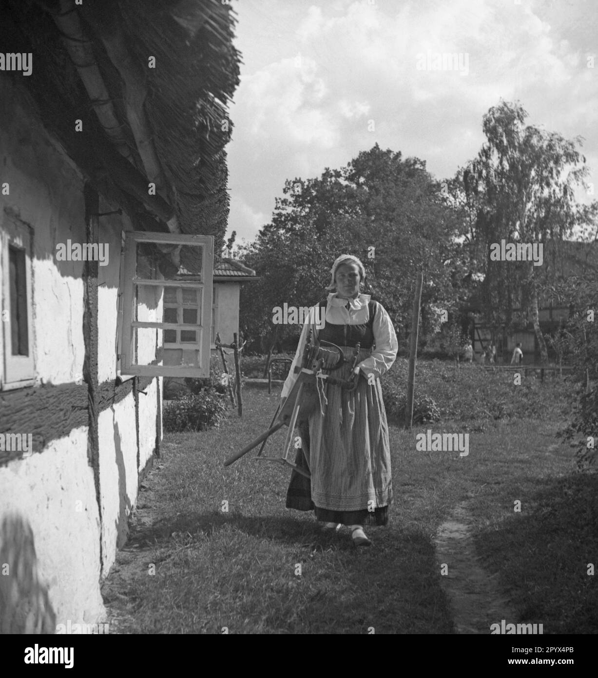 A woman carrying a spinning wheel outdoors in a village on the Baltic ...
