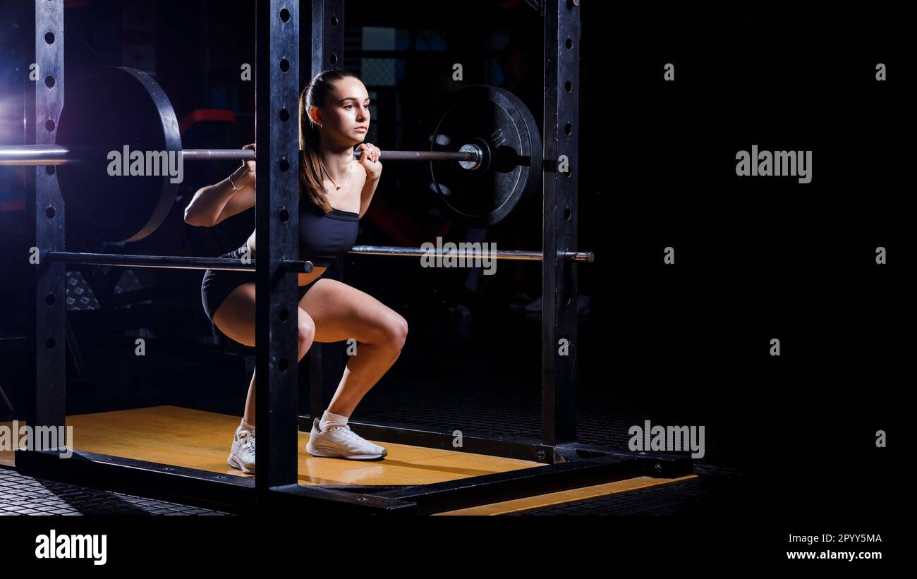Sporty young woman squatting with barbell in gym against black ...