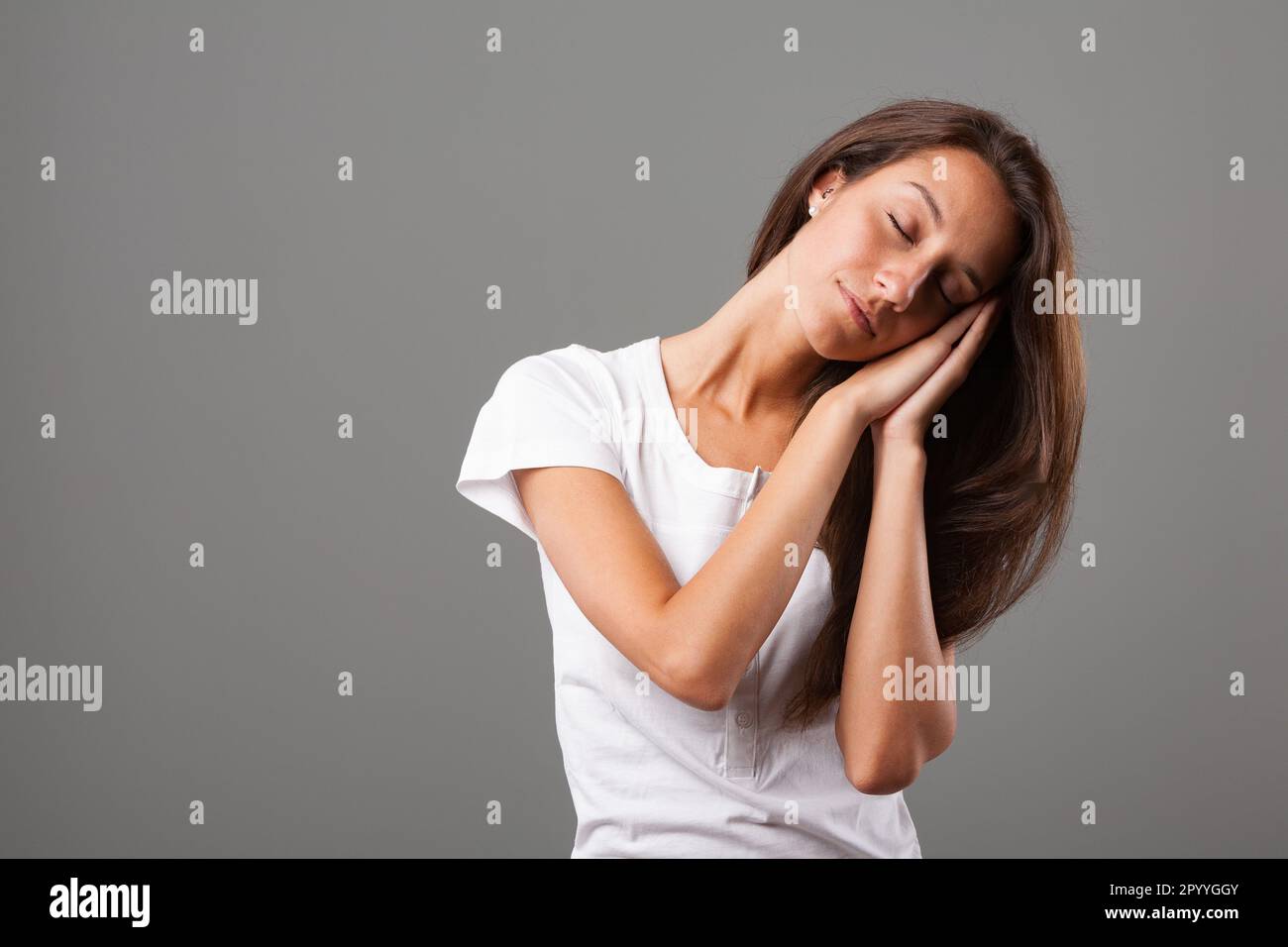 Beautiful young woman with long hair mimics sleeping gesture, hands joined, pretending to sleep ...