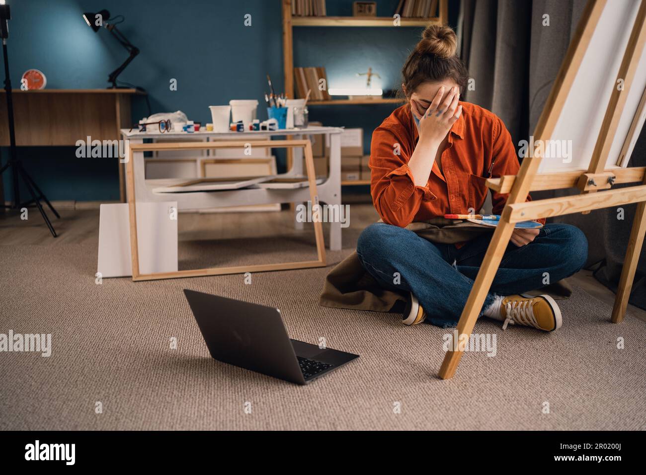 A sad artist girl in her modern studio clutched her head while sitting ...