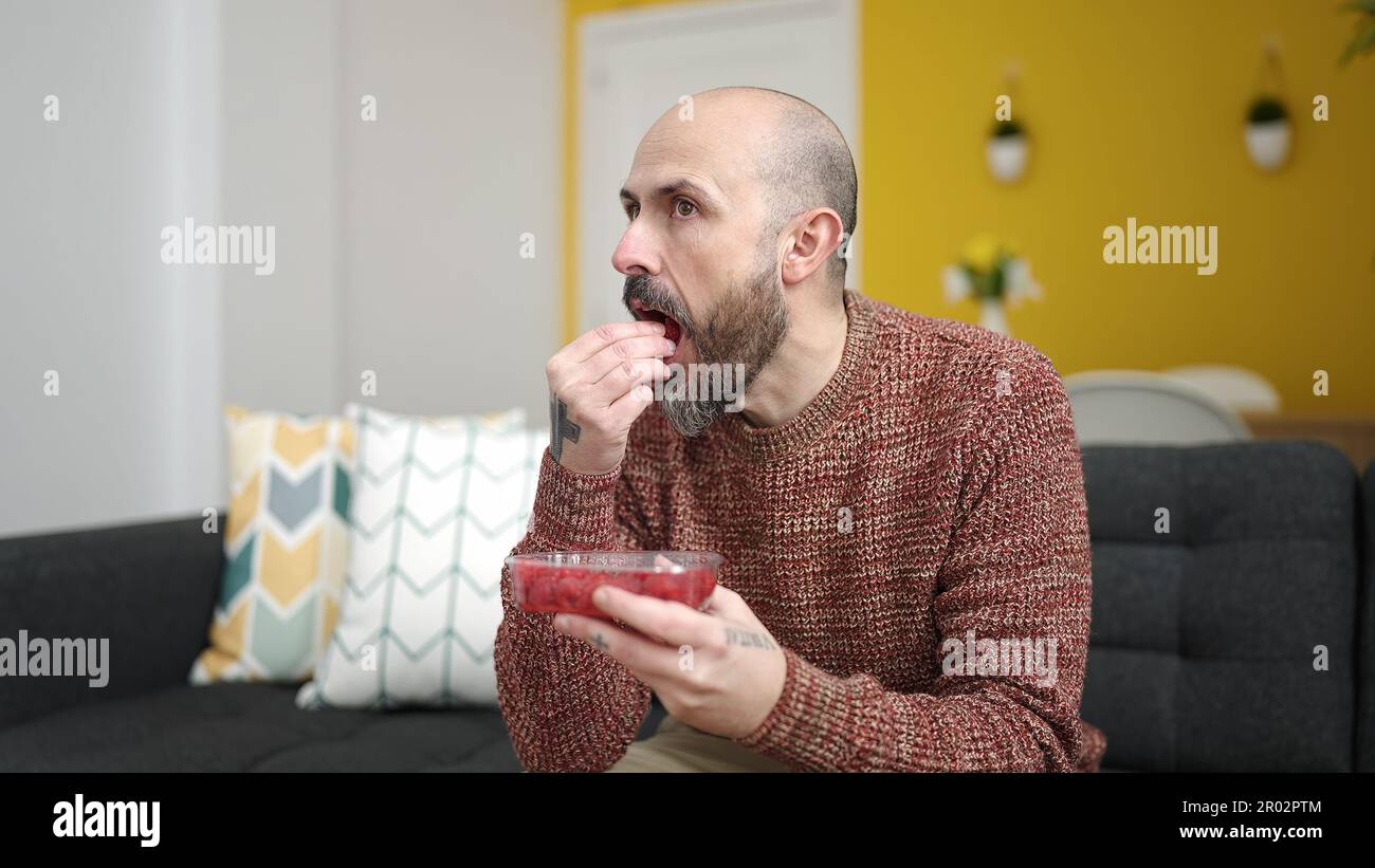 Young bald man eating raspberries sitting on sofa at home Stock Photo ...