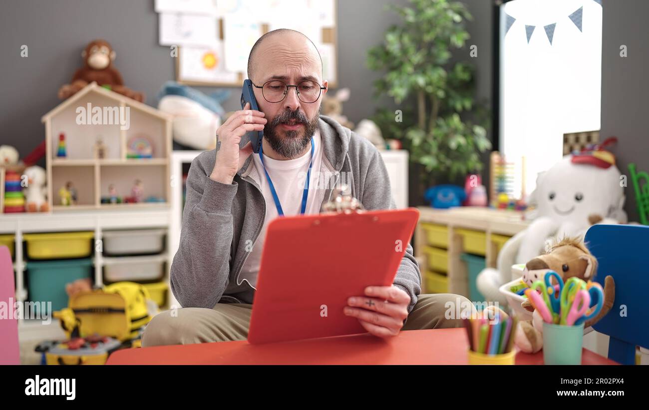 Young bald man preschool teacher reading document speaking on the phone ...