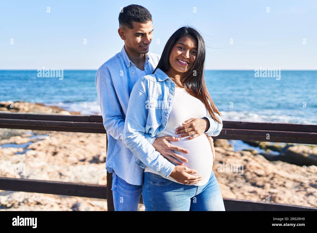Young latin couple expecting baby hugging each other standing at seaside Stock Photo - Alamy