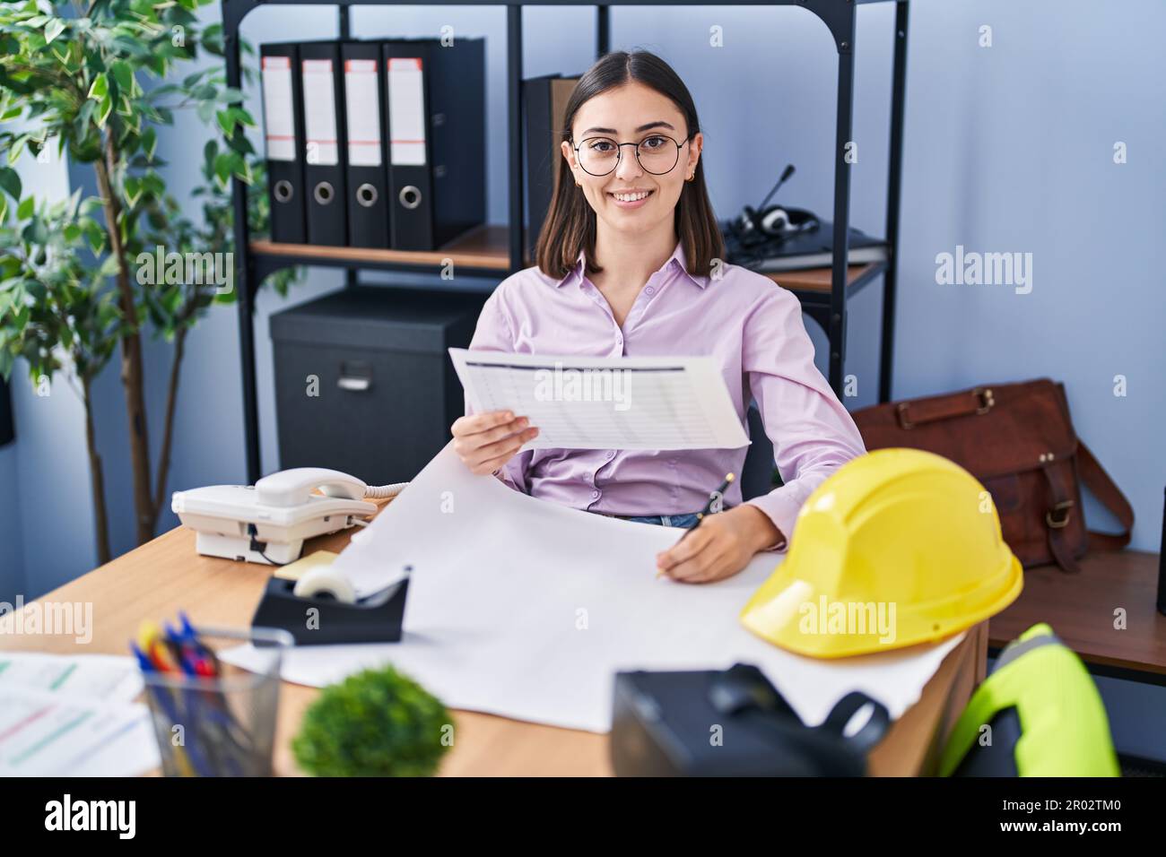 Young hispanic woman architect writing on blueprints reading document ...