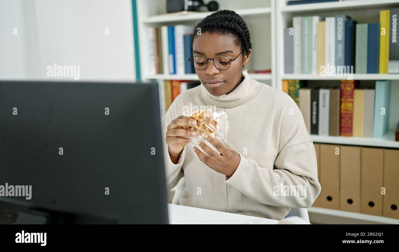 African american woman student using computer eating waffle at library ...