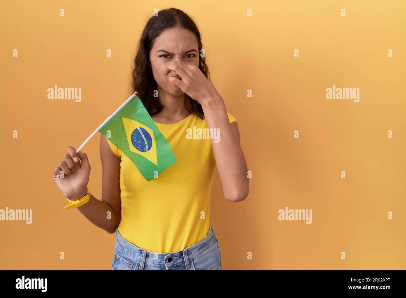 Young hispanic woman holding brazil flag smelling something stinky and ...