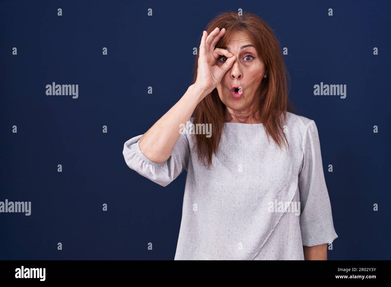 Middle age woman standing over blue background doing ok gesture shocked ...
