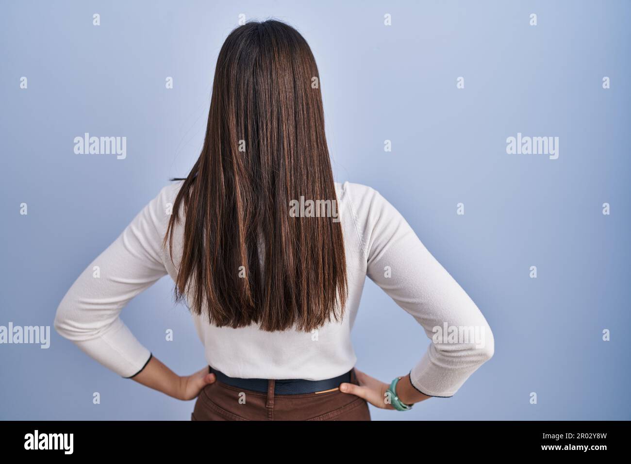 Young brunette woman standing over blue background standing backwards ...