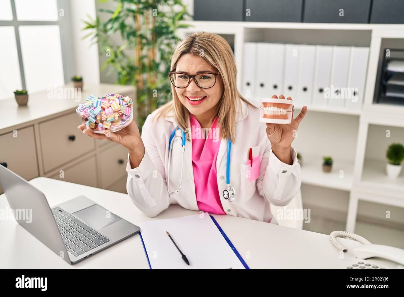 Young hispanic dentist woman holding denture and sweets clueless and ...