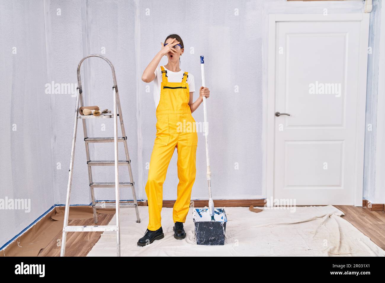 Young caucasian woman painting walls peeking in shock covering face and ...