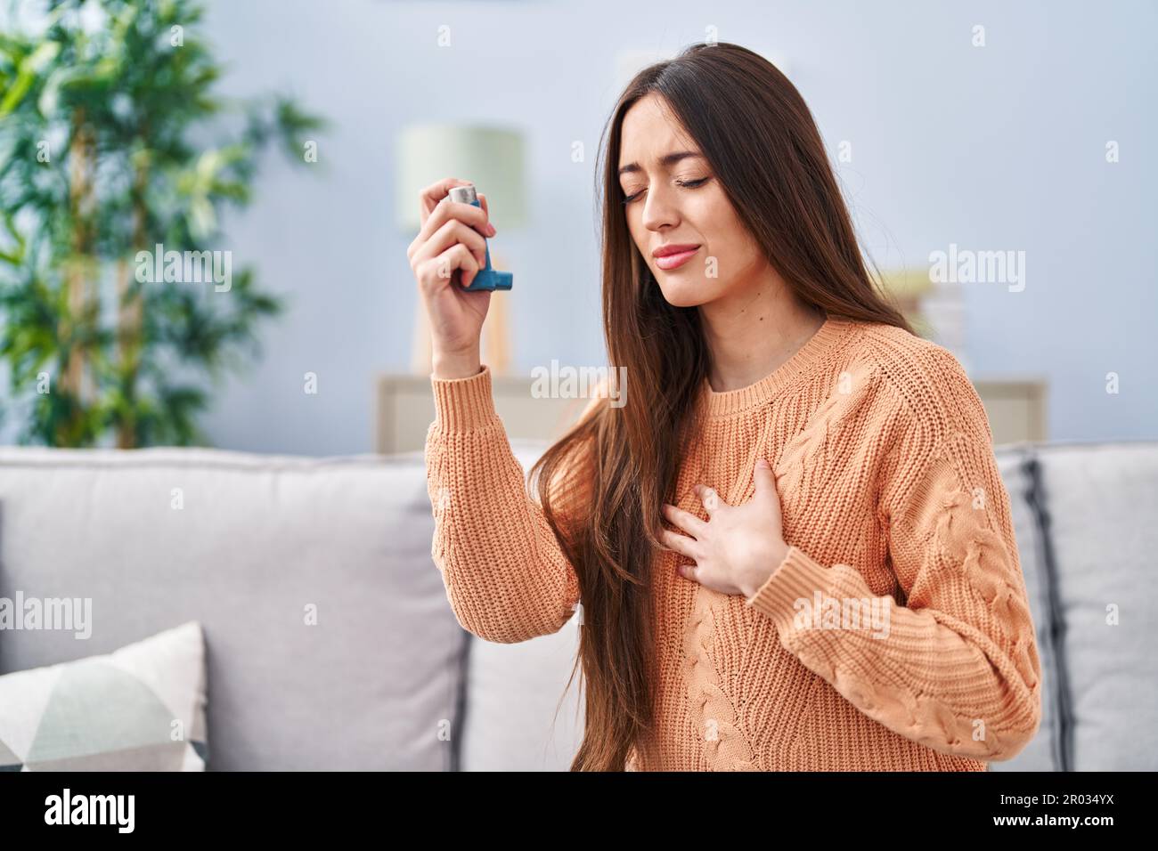 Young beautiful hispanic woman using inhaler sitting on sofa at home ...