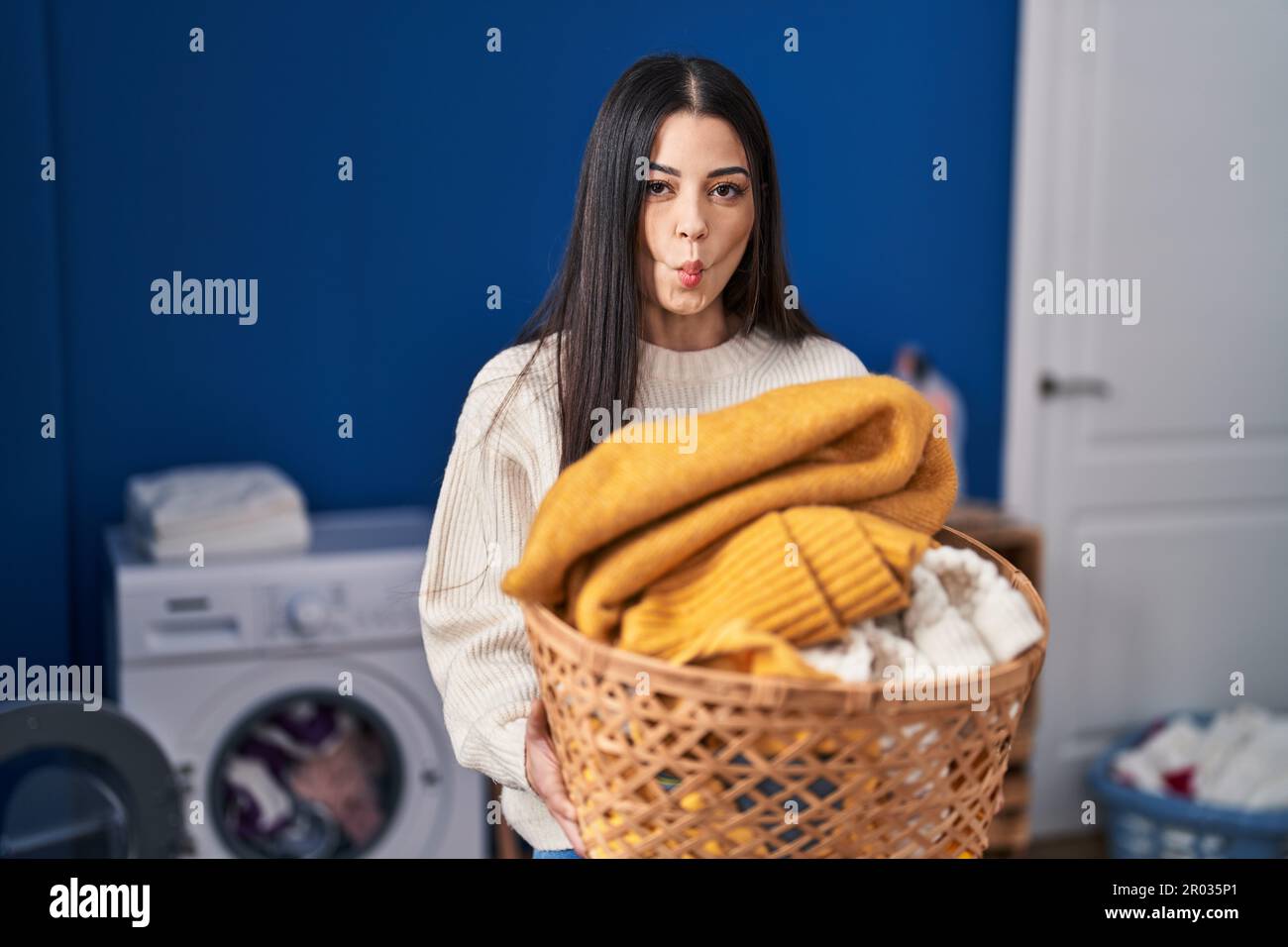 Young woman holding laundry basket making fish face with mouth and ...