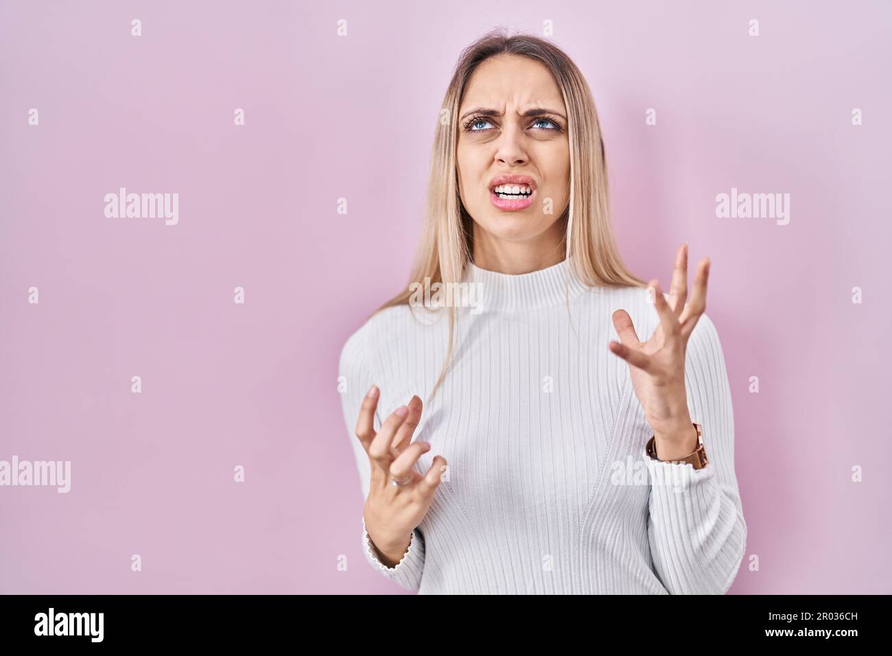 Young blonde woman wearing white sweater over pink background crazy and ...