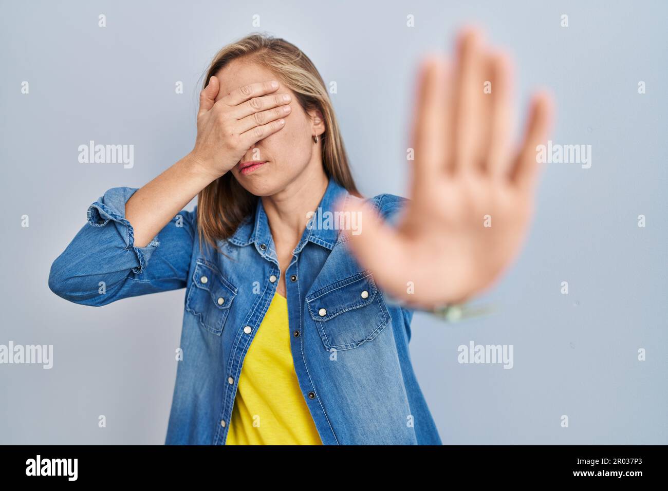 Young blonde woman standing over blue background covering eyes with ...