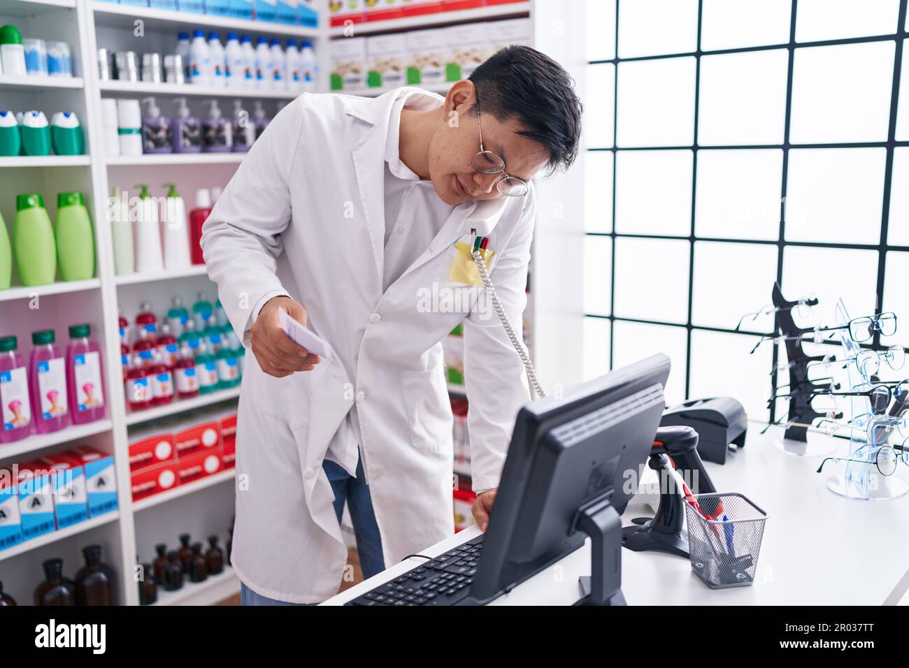 Young chinese man pharmacist talking on telephone using computer at ...