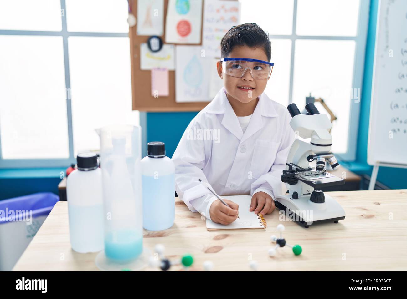 Adorable hispanic boy student using microscope writing notes at ...